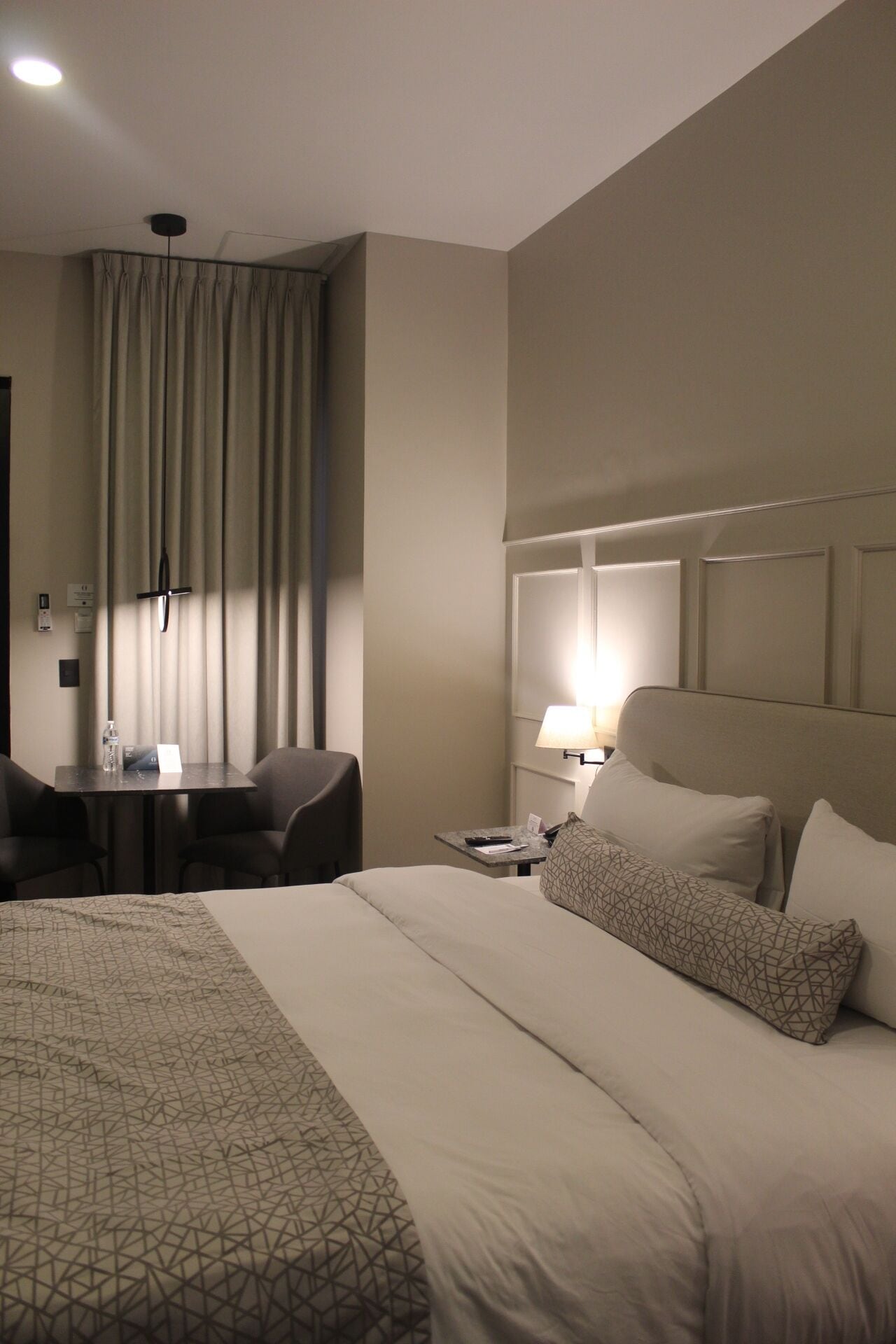 Quarto de hotel com uma cama grande, uma escrivaninha com cadeira, uma luminária e uma cortina.