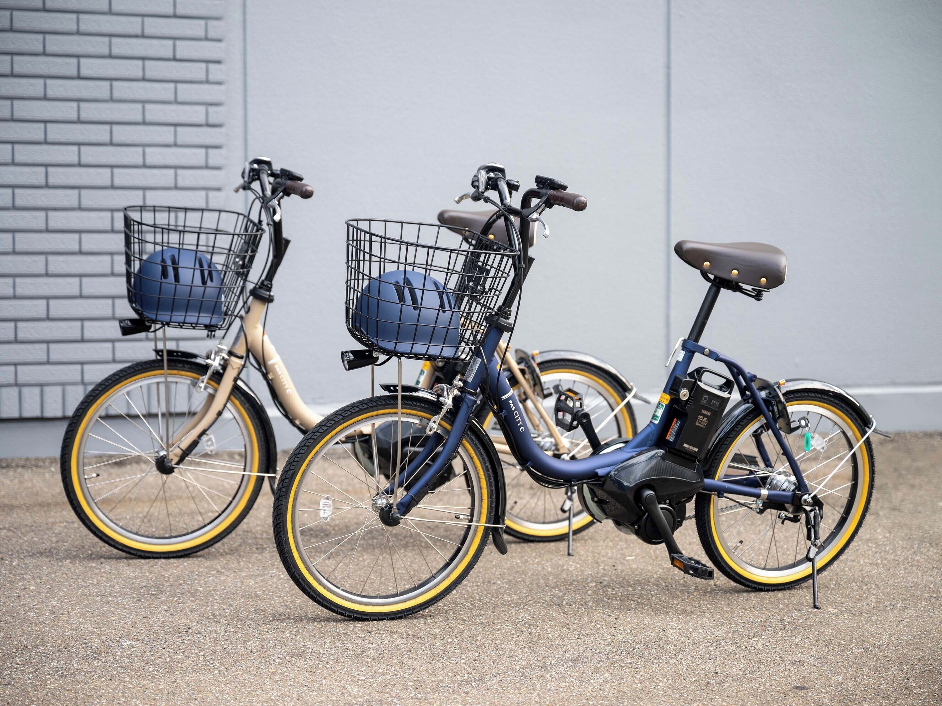 Duas bicicletas estacionadas lado a lado, com cestas na frente, com um muro de tijolos ao fundo.