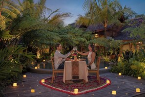 Um casal brindando com uma garrafa de vinho em uma mesa de madeira, em um espaço decorado com flores, com uma piscina e plantas tropicais ao fundo.