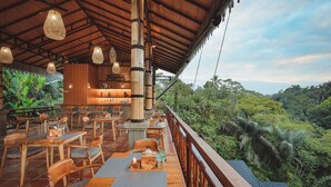 Um restaurante ao ar livre com mesas e cadeiras de madeira, um espaço de bar e vista para vegetação exuberante.