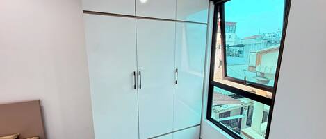 Quarto com um guarda-roupa espaçoso, uma janela com vista para edifícios e uma cama com almofada listrada.