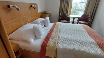 Quarto de hotel com cabeceira de madeira, cama com roupa de cama branca, duas cadeiras, uma mesinha e uma janela com cortinas.