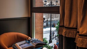 Quarto com janela, uma cadeira, uma mesa com livro e óculos, e vista para a rua com edifícios.