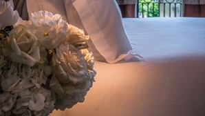 Quarto de hotel com cama, almofadas brancas, abajur, vaso de flores e vista para um edifício pela janela.