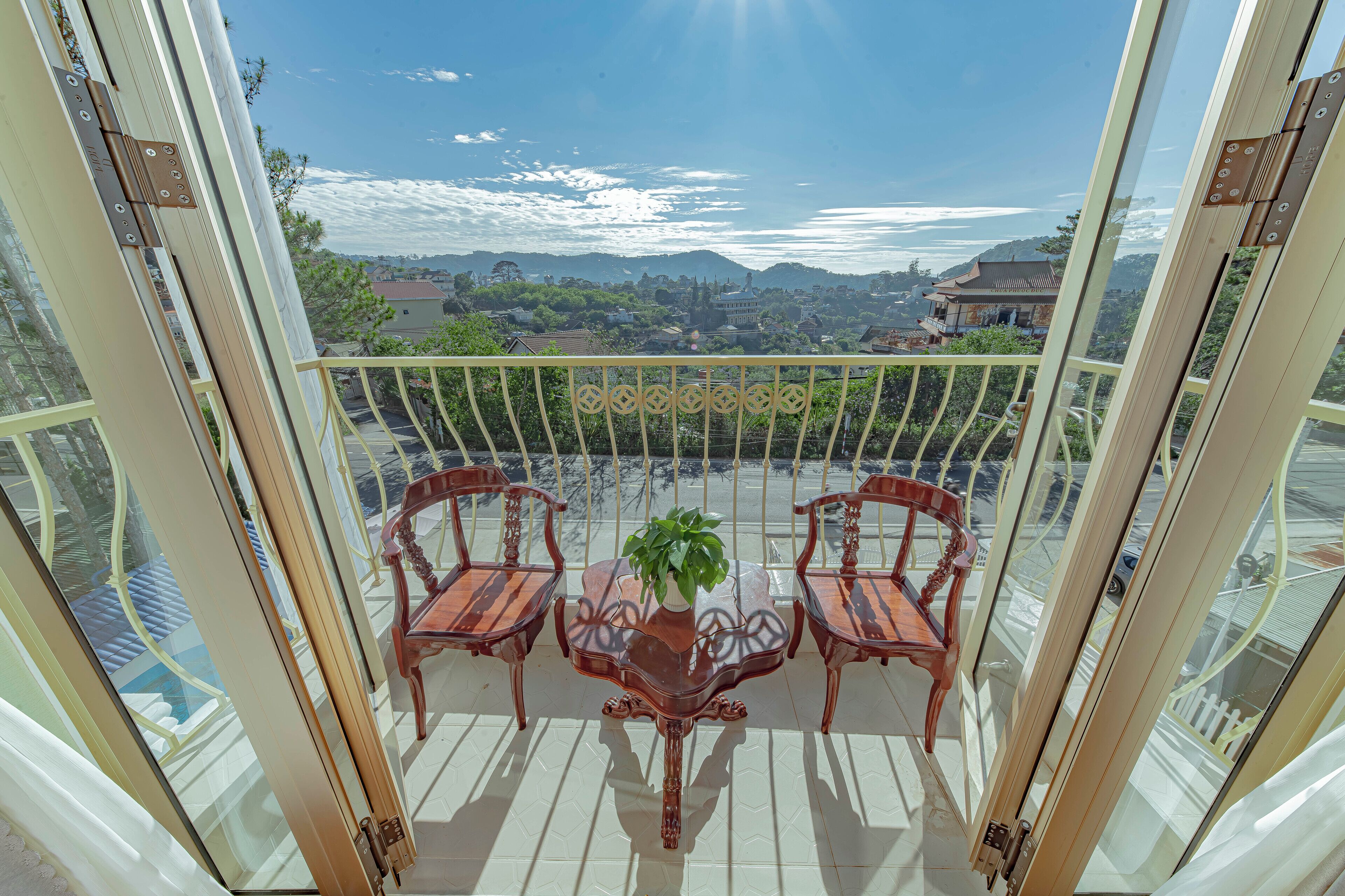Varanda com duas cadeiras de madeira e uma mesa, um vaso de planta e vista para um horizonte urbano com montanhas ao fundo.