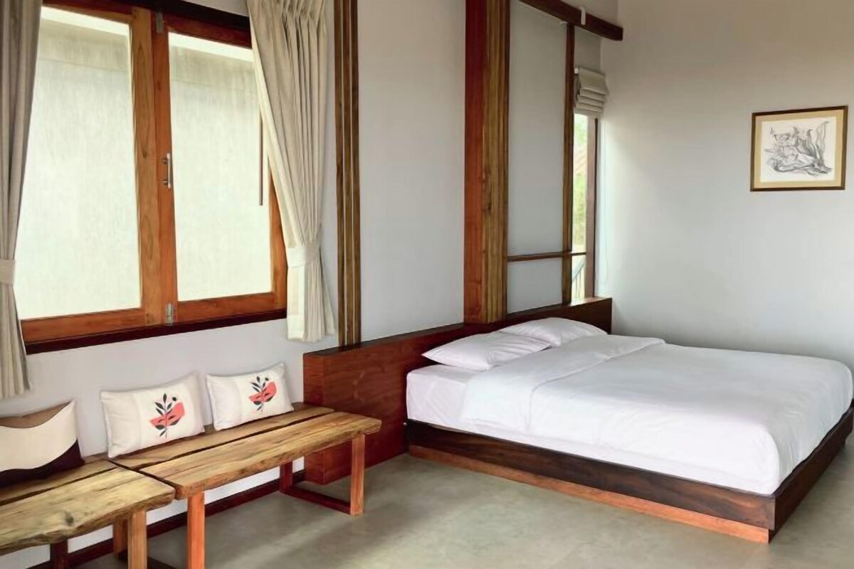 Quarto com estrutura de cama de madeira, um banco e janela com cortinas.
