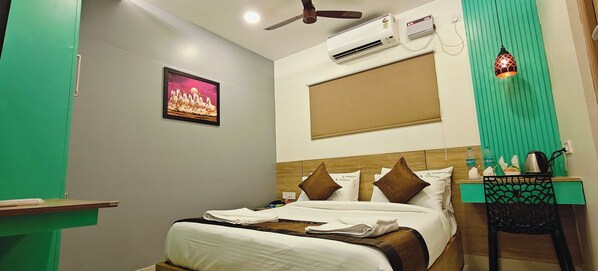 Quarto de hotel com cama, mesa de cabeceira, escrivaninha, cadeira, ar condicionado, ventoinha no teto, quadro na parede e janela com persianas.