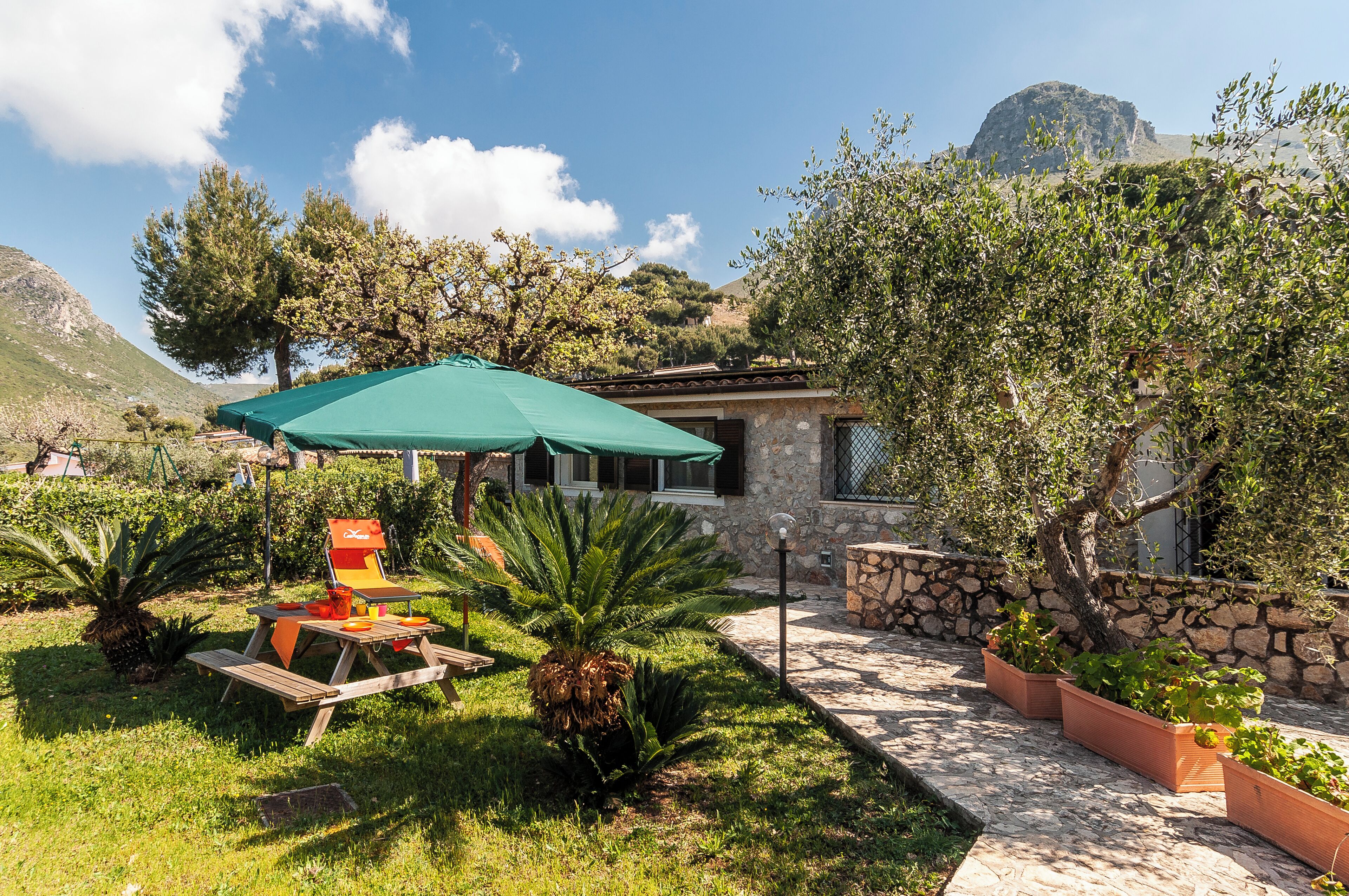 Uma casa de pedra com um guarda-sol verde, área de estar ao ar livre e um jardim com plantas e uma palmeira.