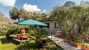Uma casa de pedra com um guarda-sol verde, área de estar ao ar livre e um jardim com plantas e uma palmeira.