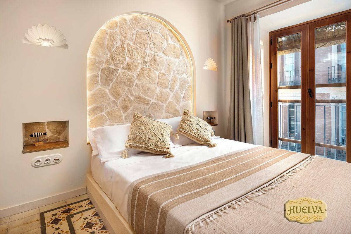 Quarto com cabeceira de pedra, cama com roupa de cama listrada, pequena prateleira com decoração de peixe e vista para o exterior por uma porta de vidro.
