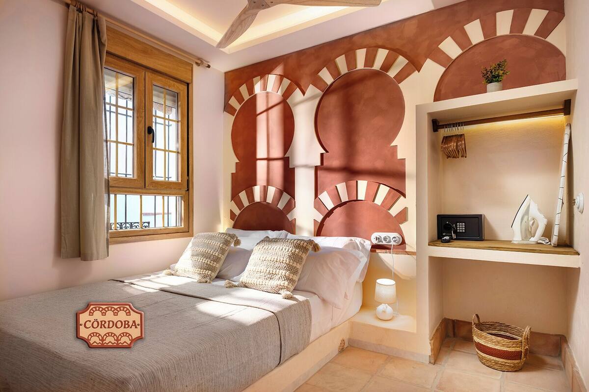Quarto com cama, mesa de cabeceira, luminária e prateleira com ferro de passar e rádio.