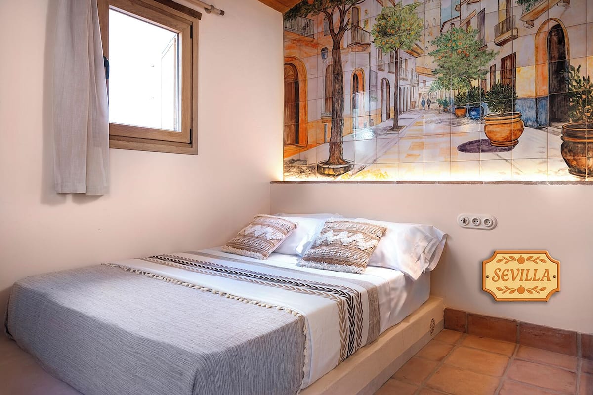 Quarto com cama, janela, quadro na parede e placa com a palavra 'SEVILLA'.