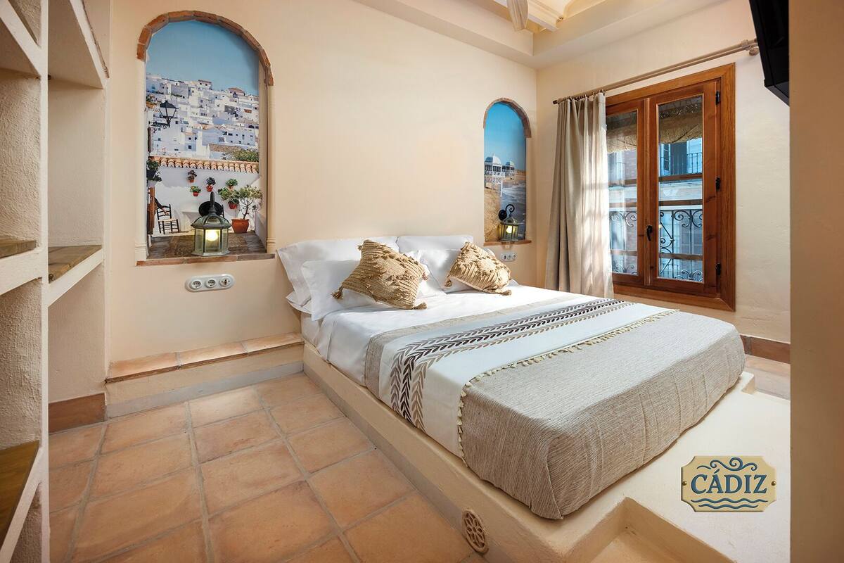 Quarto com cama, duas janelas com vista, porta de madeira e chão de azulejos.