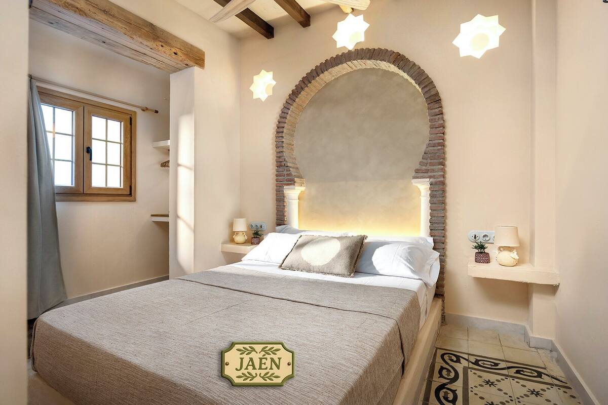 Quarto com cama, mesa de cabeceira, luminária na parede, espelho decorativo e uma pequena prateleira com uma planta.