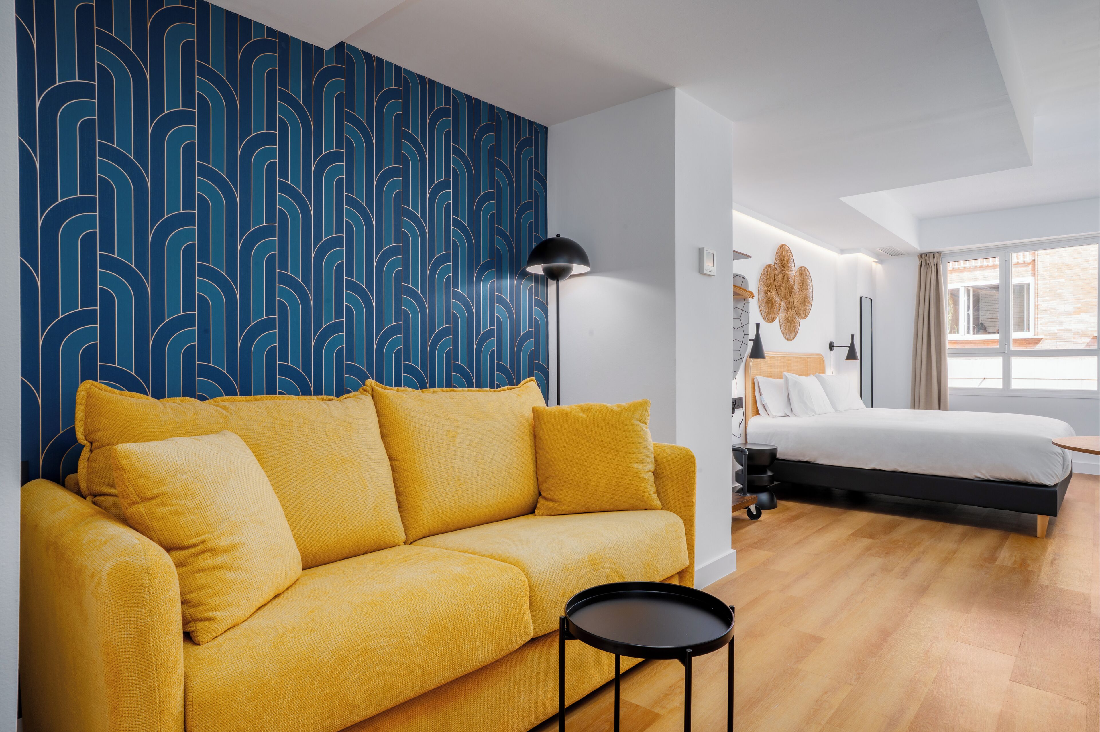 Quarto de hotel moderno com um sofá amarelo, uma cama, uma mesa lateral redonda preta e uma parede com padrão azul.