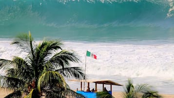 Uma cena de praia com uma onda grande, uma bandeira e uma pequena construção com pessoas.