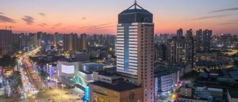 Paisagem urbana ao entardecer, com um prédio alto em destaque, apresentando um design arquitetônico único, com topo pontiagudo e uma série de janelas horizontais.