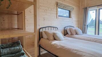 Quarto de cabana de madeira com duas camas, uma prateleira com toalhas e uma janela com cortinas.