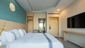 Quarto de hotel com uma cama grande, uma TV na parede, casa de banho visível através de uma porta aberta e uma pequena mesa com um vaso de flores.