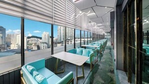 Um interior moderno com fileiras de assentos e mesas azul-turquesa, grandes janelas com vista para a cidade e um teto com luminárias lineares.