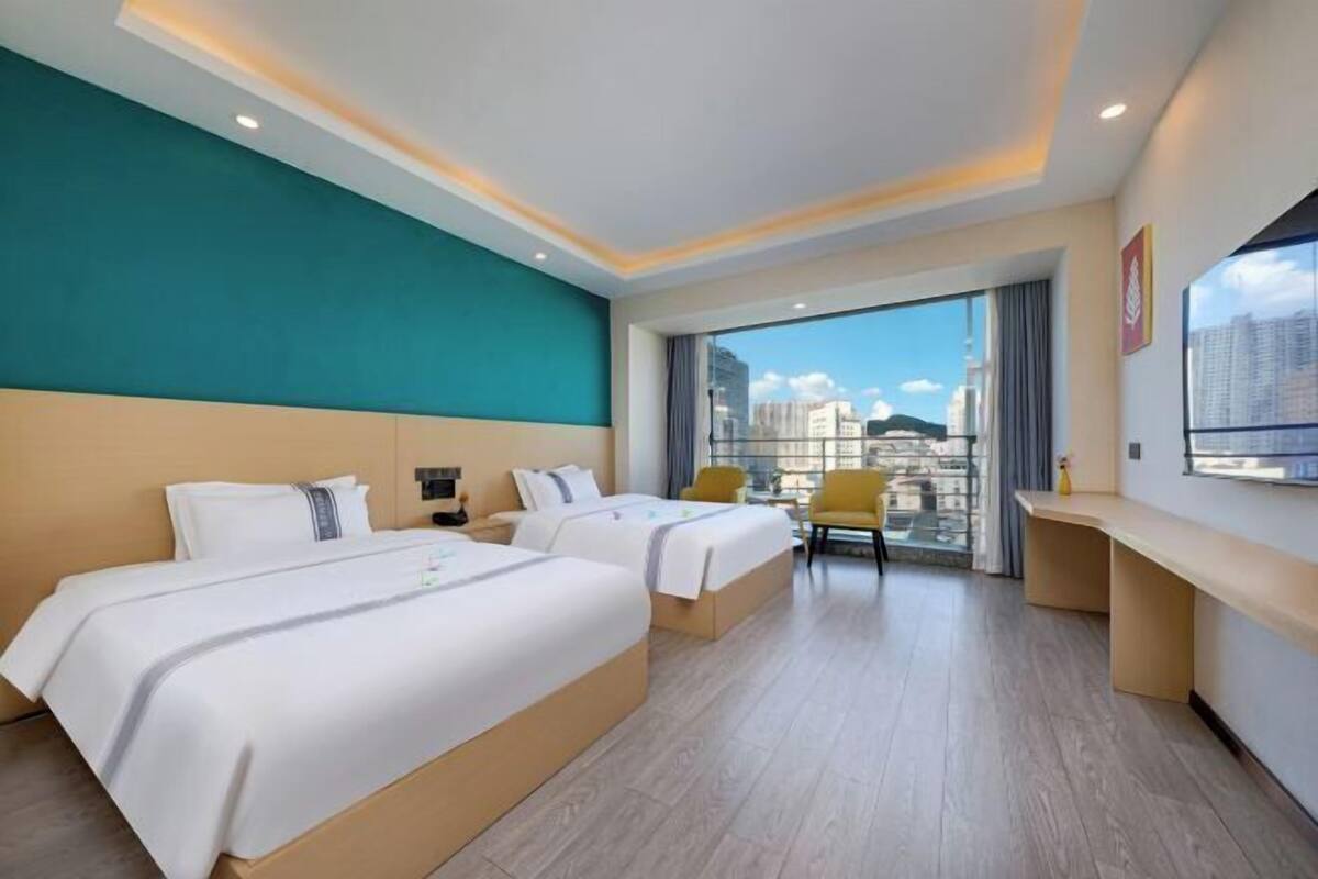 Quarto de hotel com duas camas, uma escrivaninha, uma cadeira e uma janela grande com vista para a cidade.