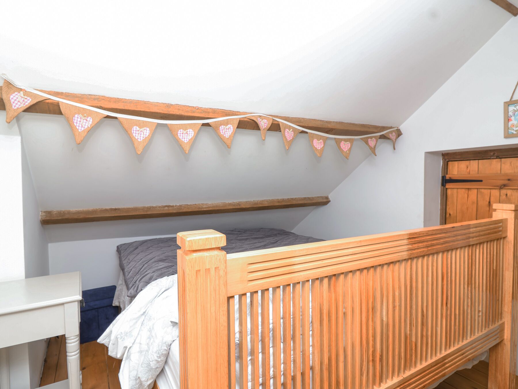 Quarto com estrutura de cama de madeira, mesa de cabeceira branca e teto inclinado decorado com guirlanda em formato de coração.