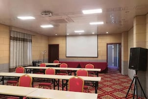 Sala de conferência com mesas, cadeiras, tela de projetor e piso coberto de carpete.