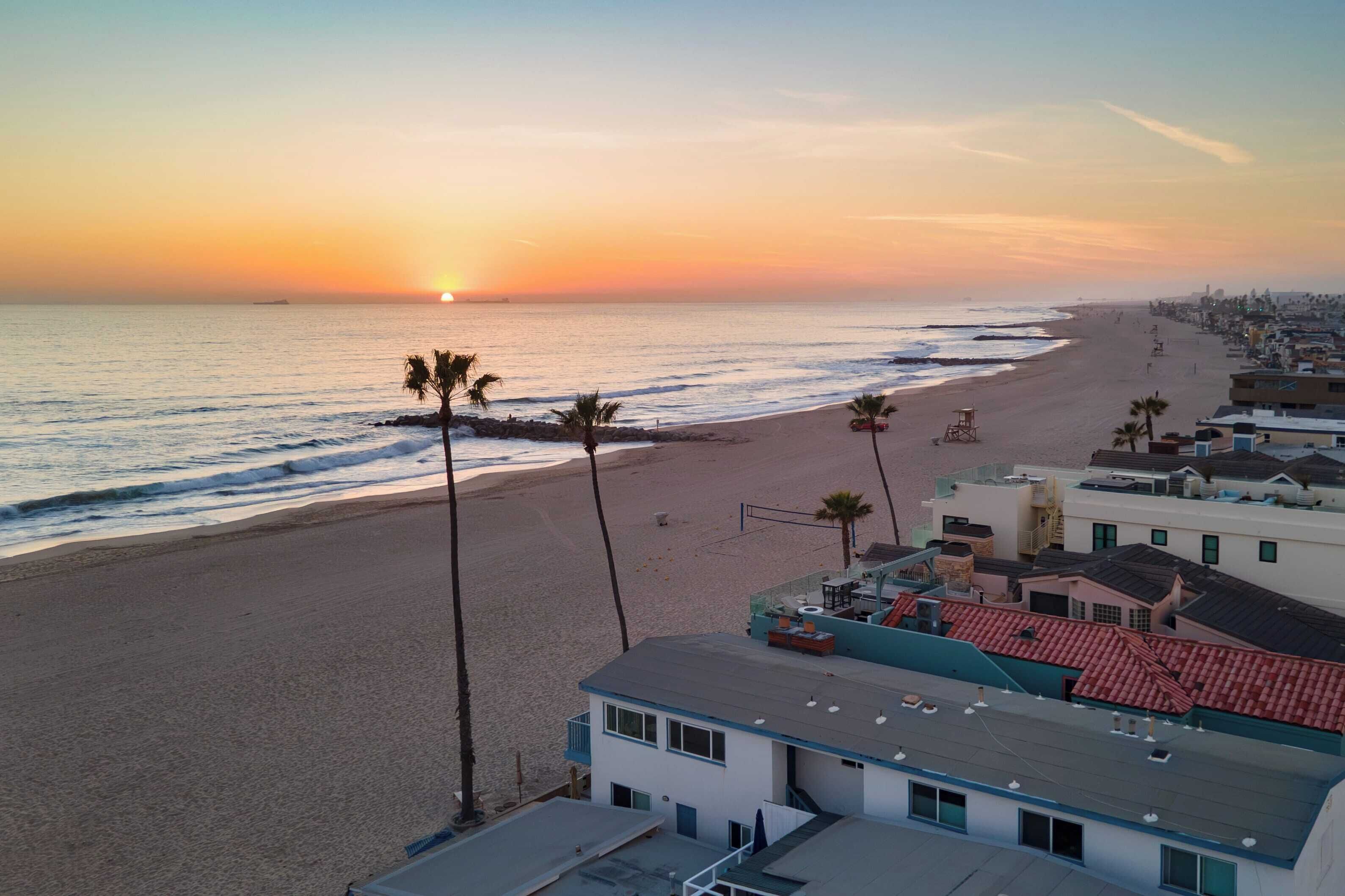 Uma praia com um pôr do sol, palmeiras e edifícios com telhados vermelhos.