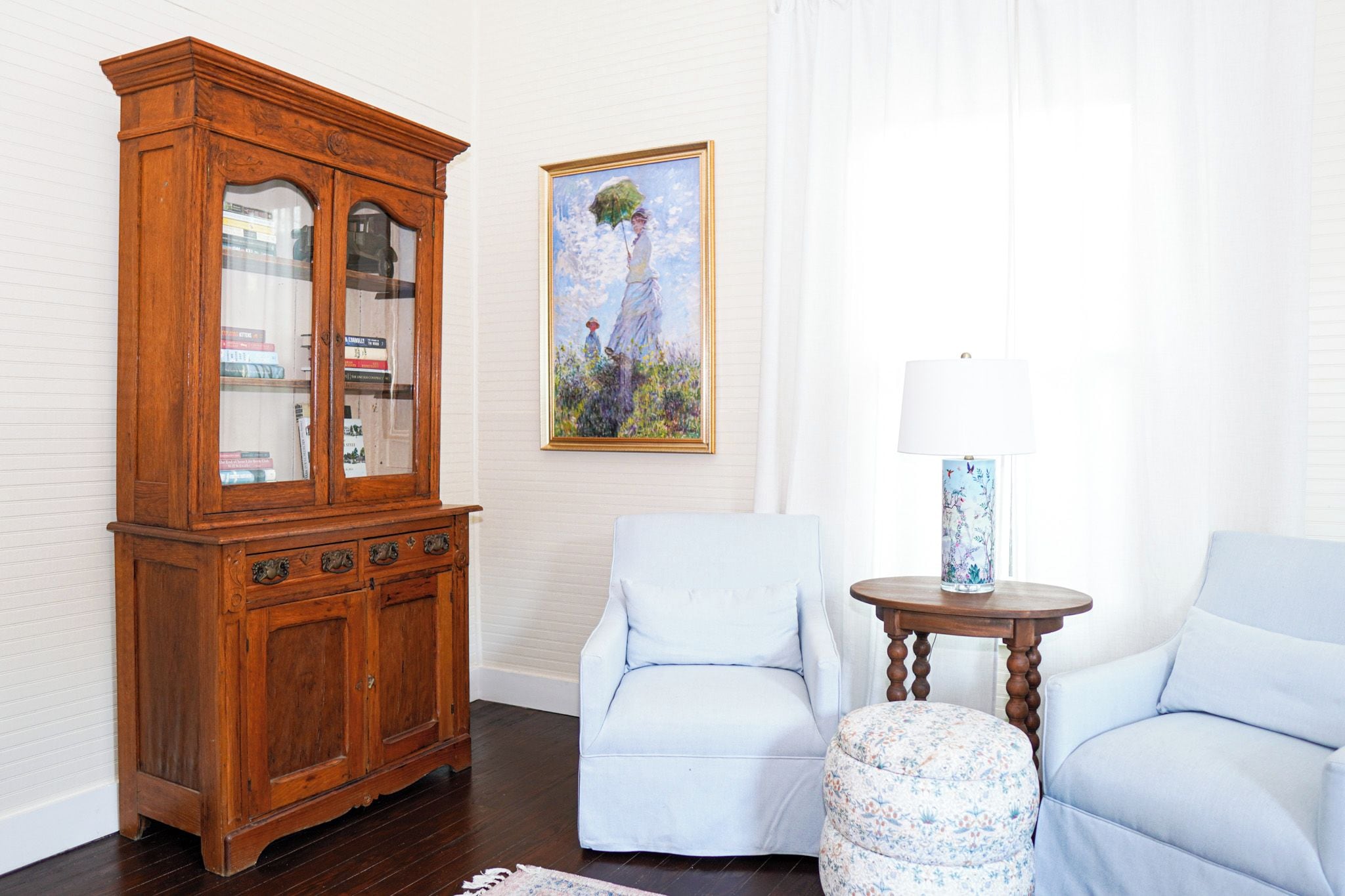 Quarto com armário de madeira, duas poltronas estofadas, uma pequena mesa redonda com abajur e um quadro pendurado na parede.