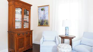 Quarto com armário de madeira, duas poltronas estofadas, uma pequena mesa redonda com abajur e um quadro pendurado na parede.