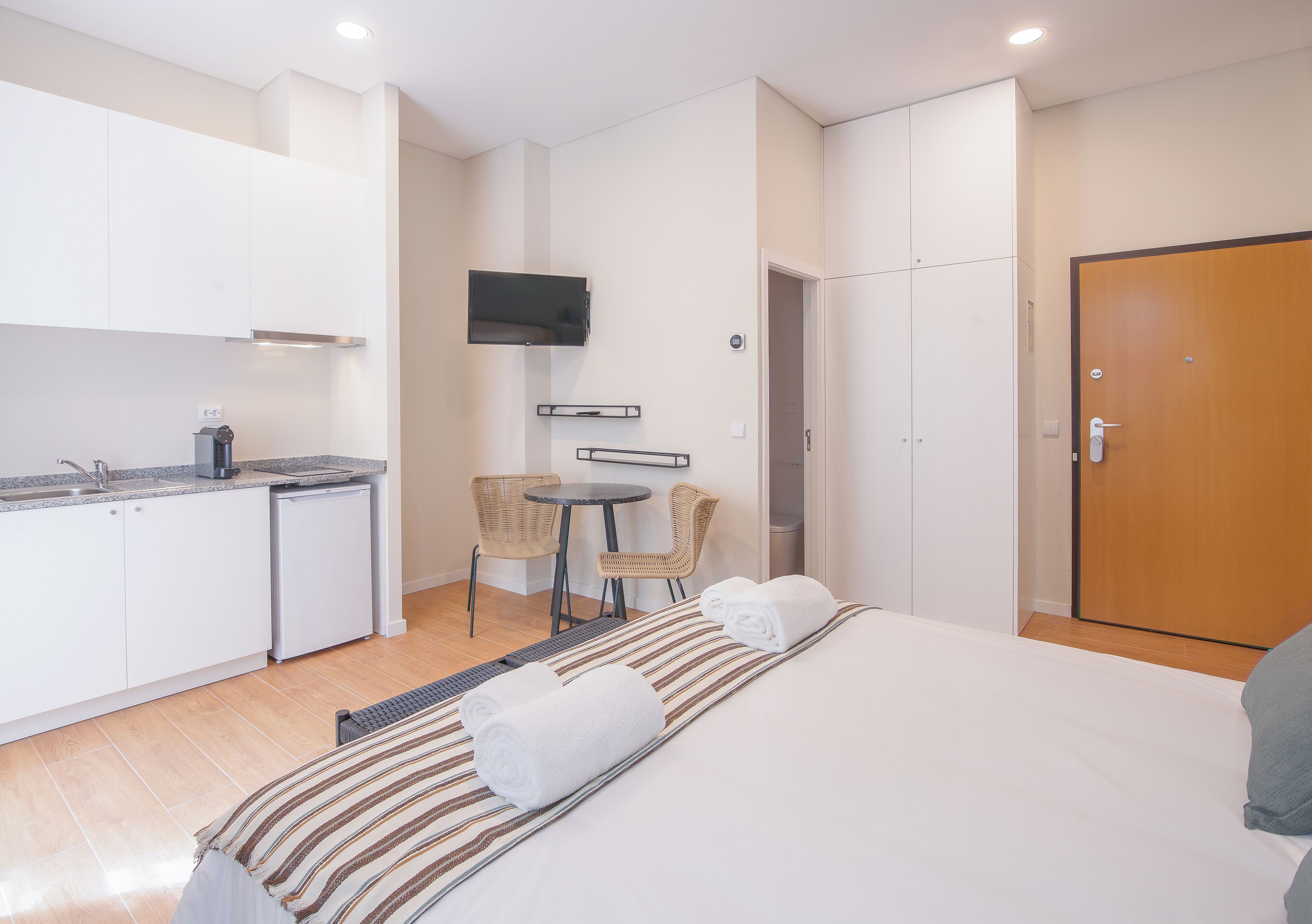 Quarto de hotel com cama, cozinha compacta, televisão, uma cadeira e porta de madeira.