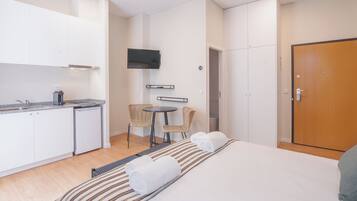 Quarto de hotel com cama, cozinha compacta, televisão, uma cadeira e porta de madeira.