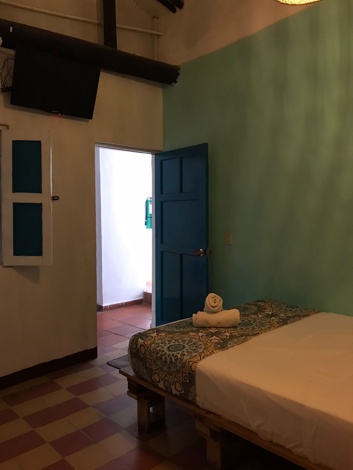Quarto com cama, um banquinho, porta azul e televisão fixada na parede.
