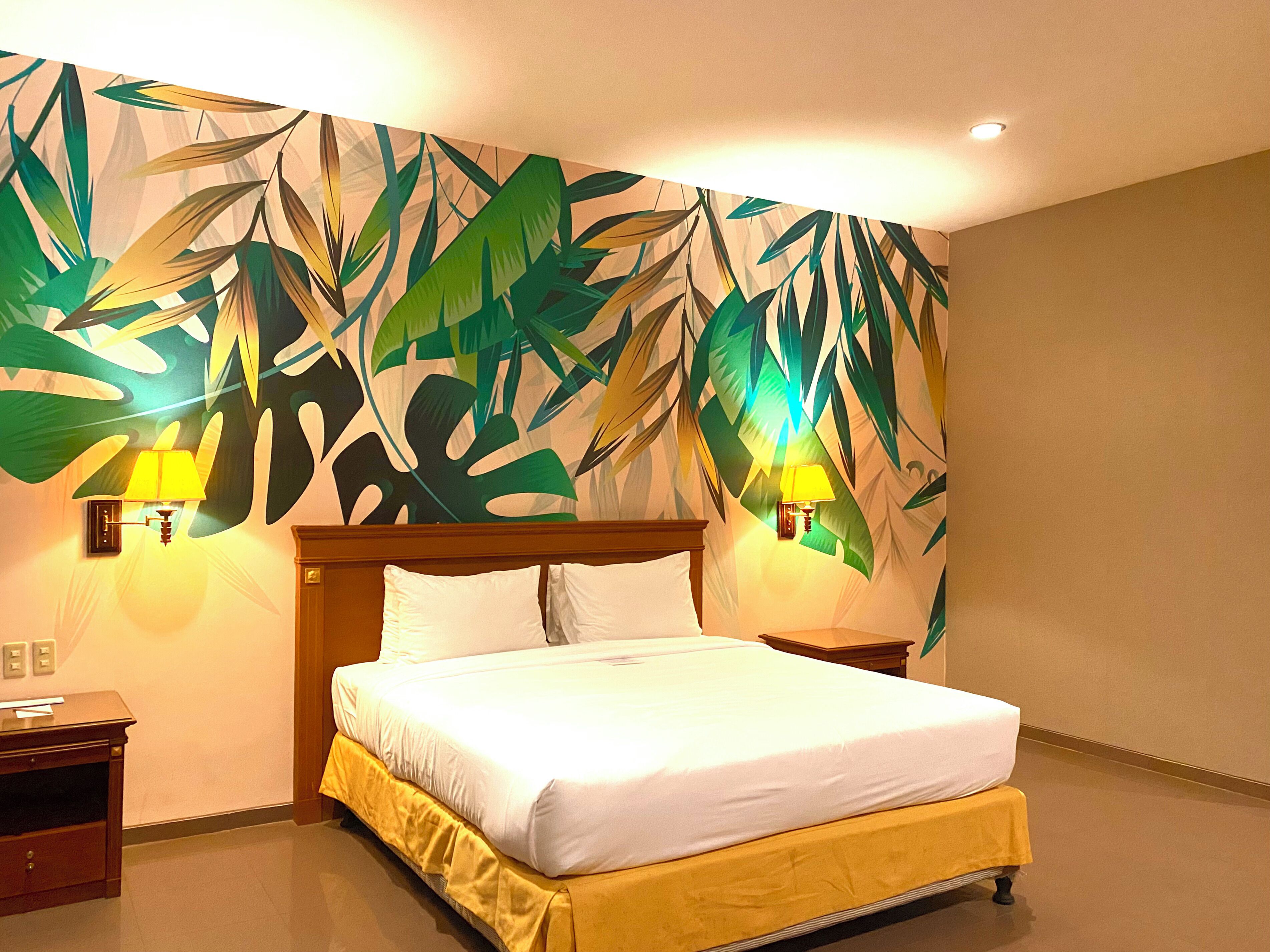 Quarto de hotel com uma cama grande, duas luminárias de cabeceira e um papel de parede com padrão de folhagem tropical.