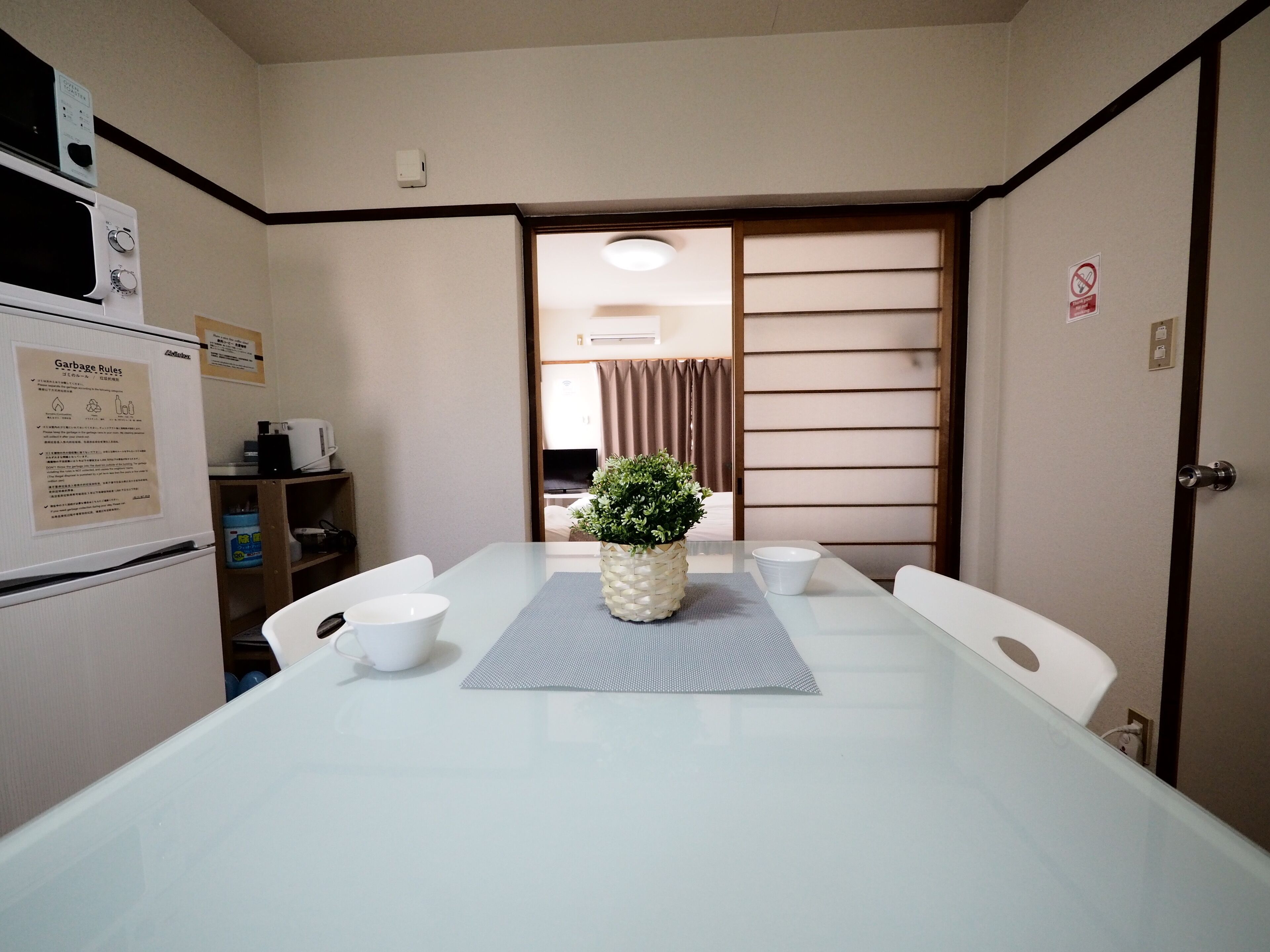 Quarto de hotel com mesa de jantar, cadeiras, planta em vaso, micro-ondas, frigorífico e porta deslizante para um banheiro.