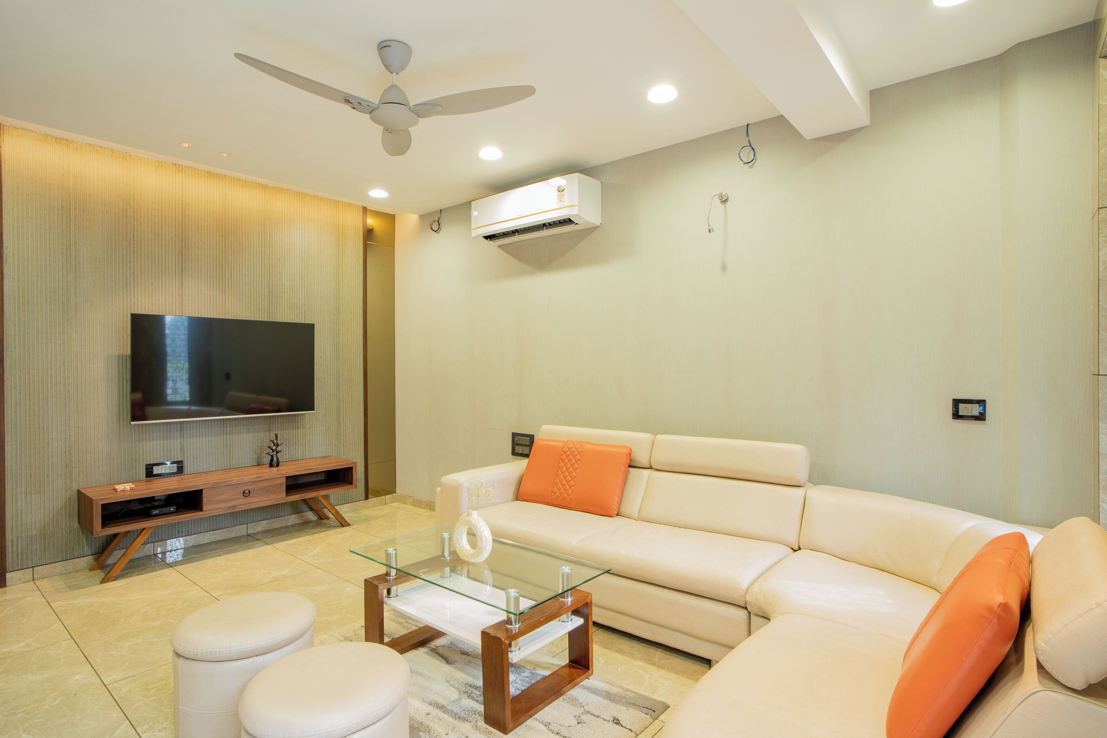 Uma sala de estar moderna com uma TV de tela plana, uma mesa de centro de vidro, um sofá bege com almofadas laranja, um ventilador de teto e um aparelho de ar condicionado.