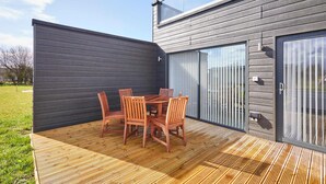 Um pátio externo com mesa e cadeiras de madeira, uma porta de correr de vidro e uma parede externa escura.