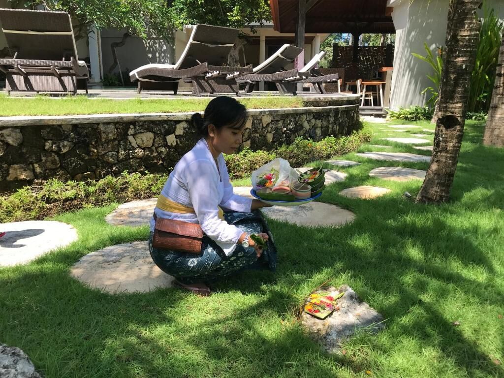 Uma pessoa sentada na grama em uma área de jardim, com cadeiras de descanso e um muro de pedra ao fundo.