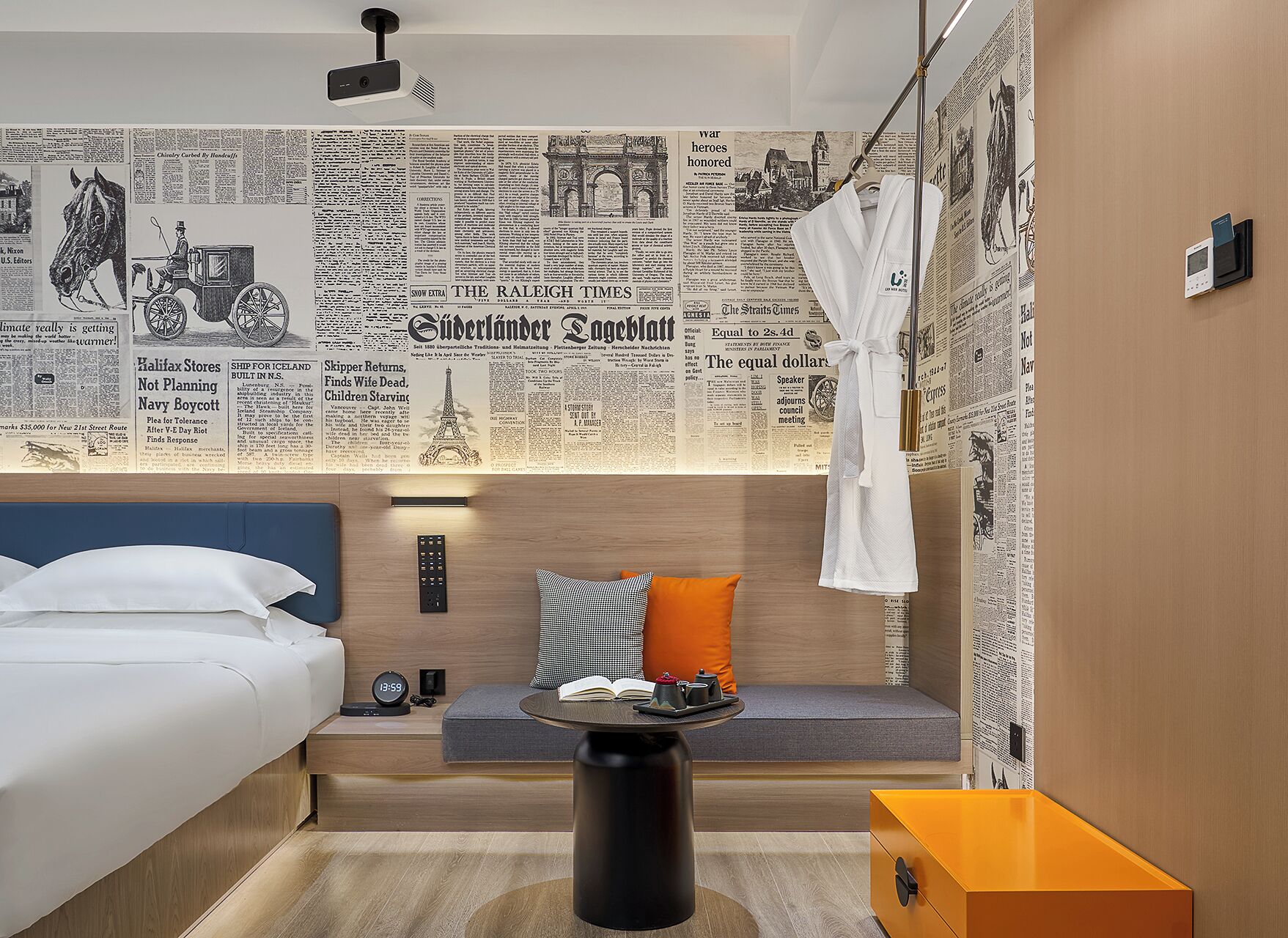 Quarto de hotel moderno com uma parede grande coberta de jornais, uma cama, uma mesa de cabeceira e um pequeno espaço de estar com um banco e almofadas.