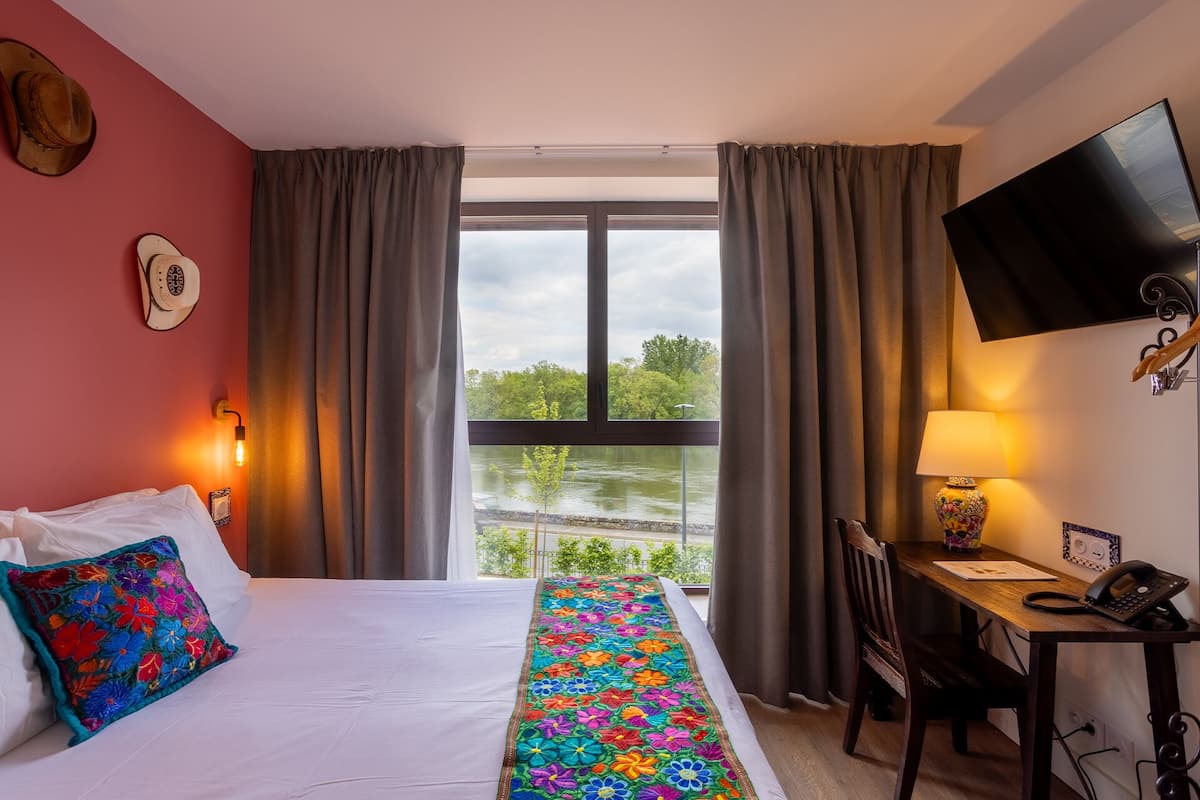 Quarto de hotel com cama, escrivaninha, cadeira, televisão e janela com cortinas.