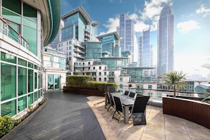 Terraço no telhado com área de jantar, rodeado por edifícios modernos e céu claro.