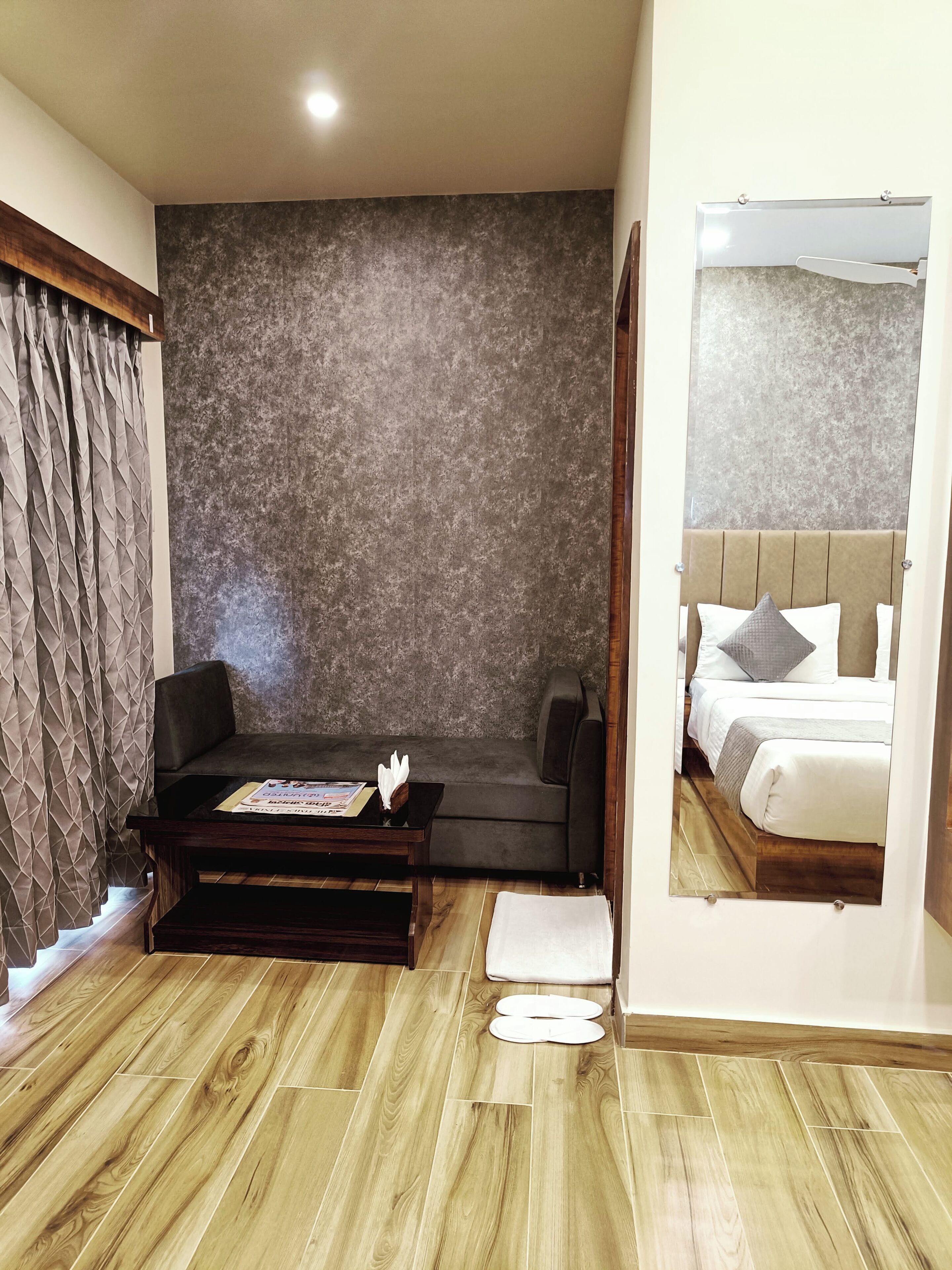 Quarto de hotel moderno com piso de madeira, sofá escuro, pequena mesa de centro e um espelho grande que reflete uma cama com roupa de cama branca e travesseiro cinza.