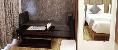 Quarto de hotel moderno com piso de madeira, sofá escuro, pequena mesa de centro e um espelho grande que reflete uma cama com roupa de cama branca e travesseiro cinza.