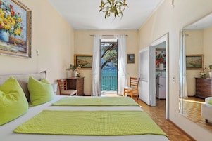 Quarto com cama, almofadas verdes, escrivaninha e vista para o mar através de uma porta.