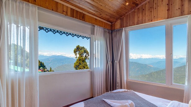 Quarto com uma cama grande, teto de madeira e vista para montanhas através de duas janelas.