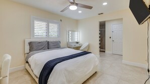 Quarto com cama, ventilador de teto, toucador com espelho e porta que dá para outro cômodo.