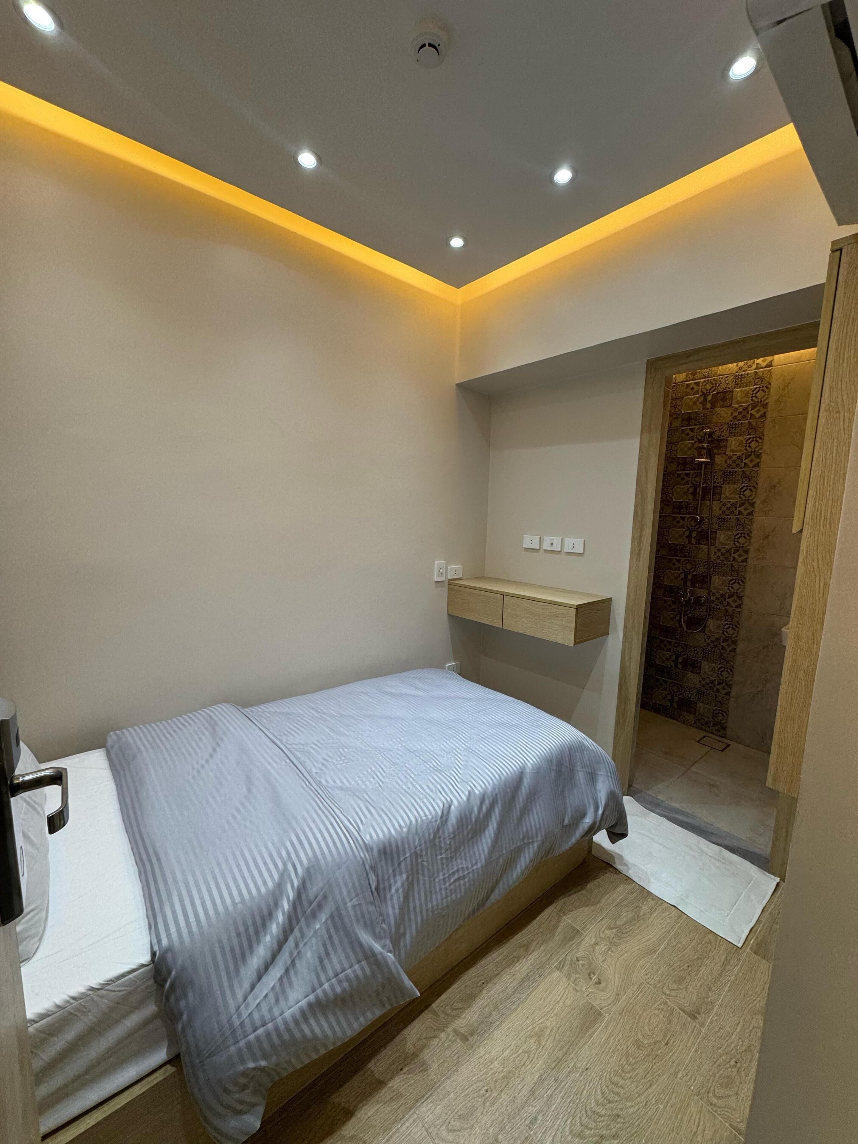 Quarto pequeno e moderno com uma cama de casal, uma mesa de cabeceira de madeira, uma estante embutida e um chuveiro com azulejos estampados.