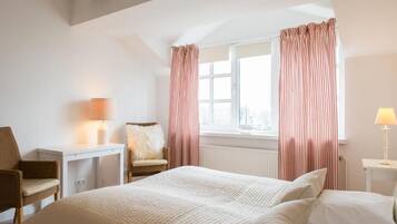 Quarto de hotel com cama, escrivaninha, cadeira e janela com cortinas.