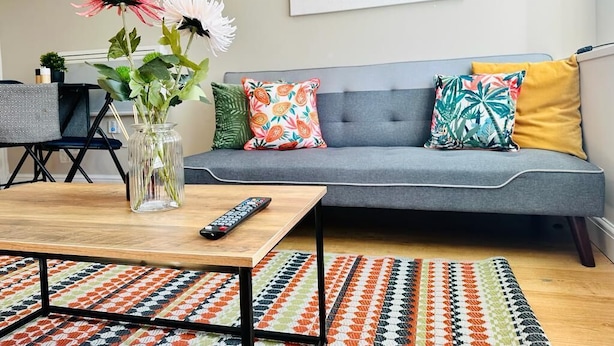 Uma sala de estar com um sofá cinza, almofadas coloridas, uma mesa de centro de madeira e um vaso com flores.