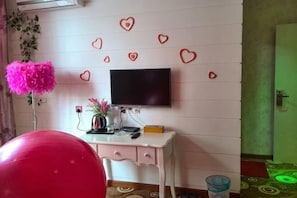 Um quarto com escrivaninha, televisão, bola de exercício rosa e decorações em formato de coração na parede.